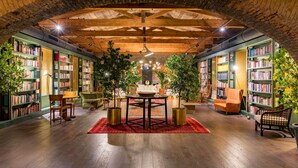 Uma biblioteca com estantes, uma mesa central, cadeiras e plantas em vasos.