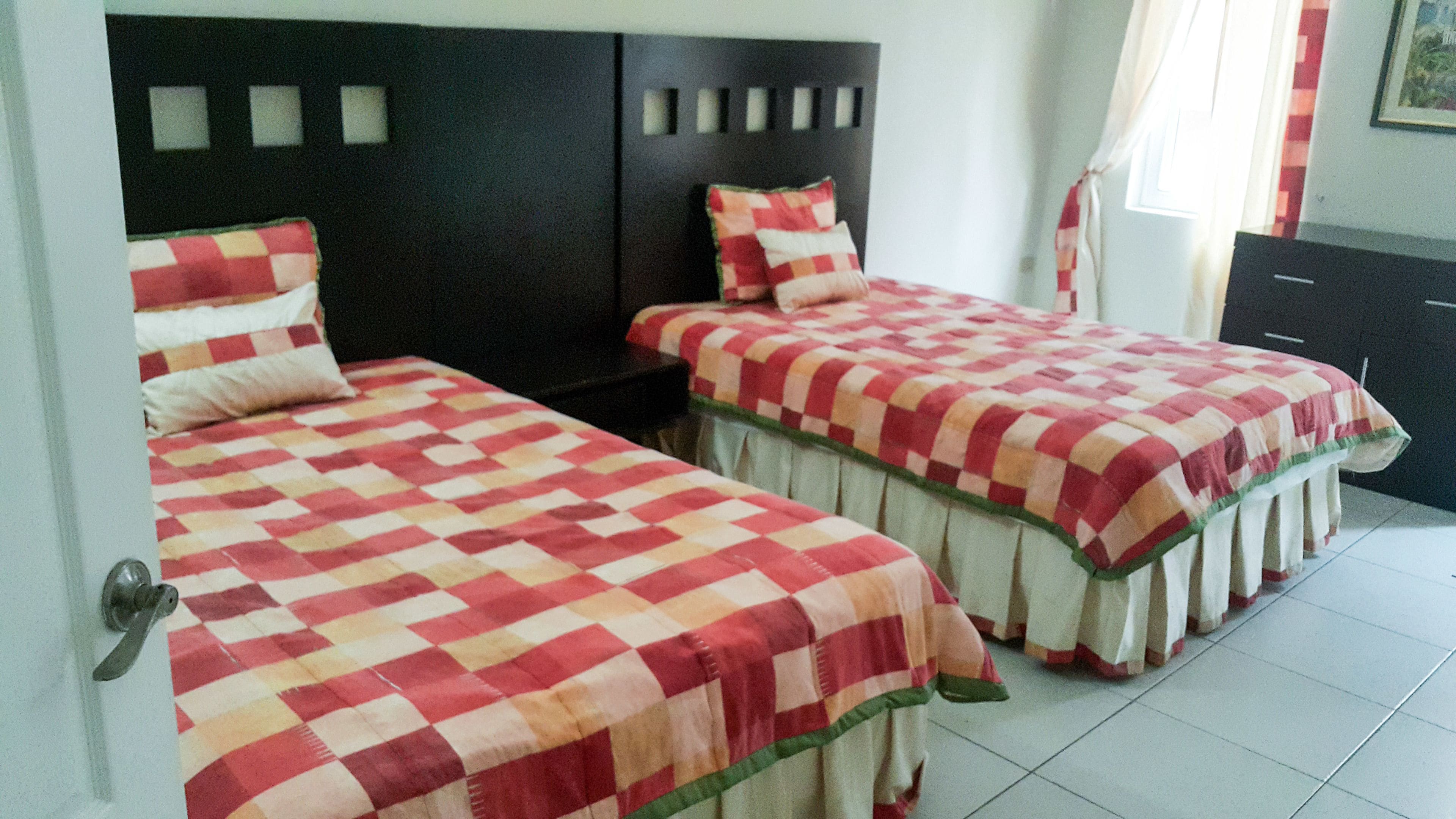 Quarto com duas camas, cada uma com colcha xadrez e almofadas. Apresenta uma cabeceira escura, uma mesinha e um armário.