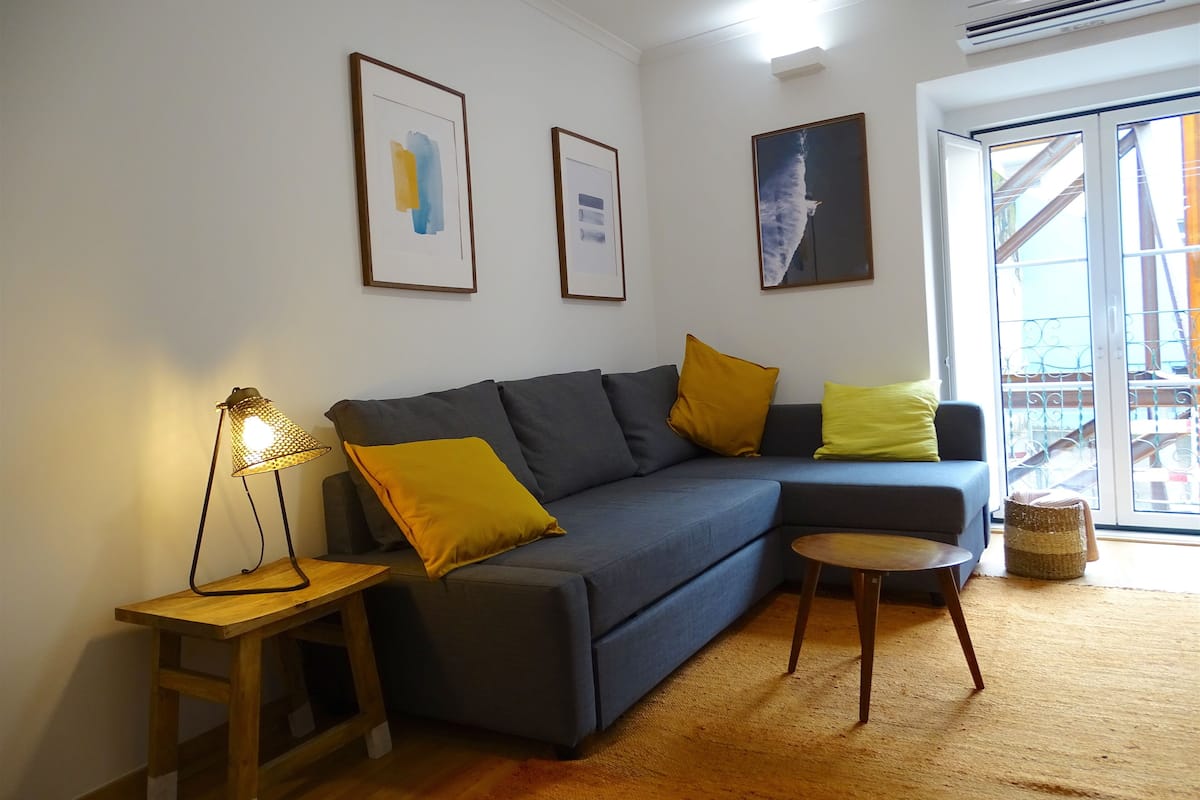 Sala de estar moderna com um sofá modular cinza, almofadas amarelas e verdes, uma pequena mesa de madeira e um abajur. Há obras de arte emolduradas na parede e uma grande janela com vista para uma varanda.