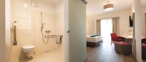 Quarto de hotel moderno com casa de banho, chuveiro, cama e uma pequena área de estar com uma cadeira e mesa.