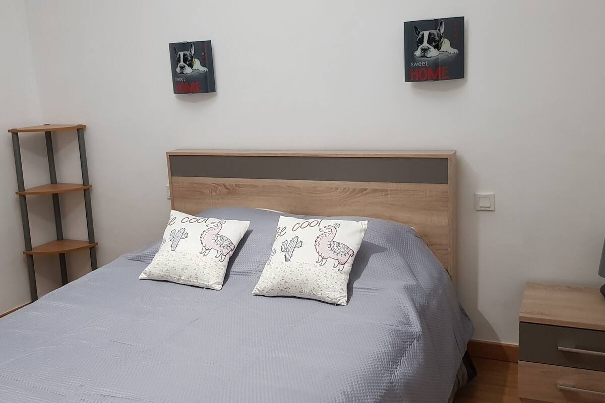 Quarto com cama, duas almofadas, mesa de cabeceira com abajur e duas imagens penduradas na parede.