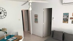 Uma sala de estar moderna com um sofá cinzento, uma área de jantar com mesa e cadeiras de madeira, um ventilador de teto e um relógio de parede.