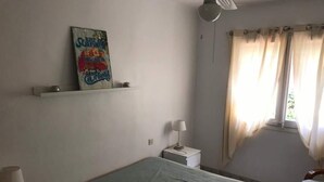 Quarto com cama, mesa de cabeceira, luminária, prateleira com pôster e janela com cortinas.