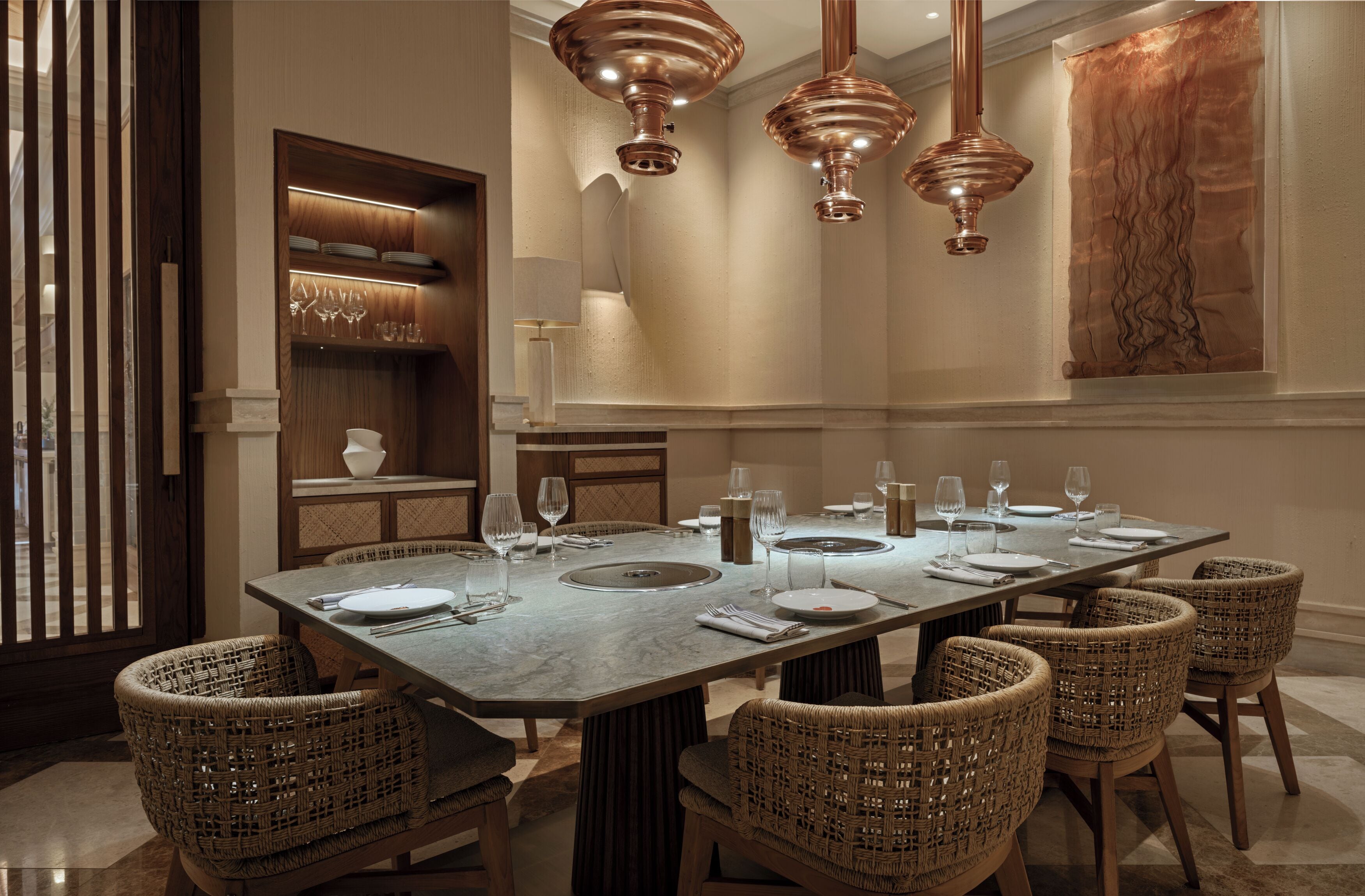 Uma área de refeições com uma mesa comprida, cadeiras de vime e luminárias pendentes de cobre.