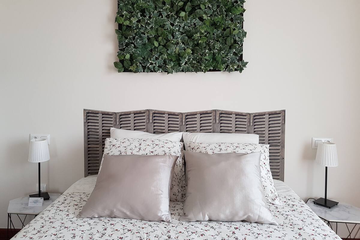 Quarto bem arrumado com uma cama, duas mesas de cabeceira e uma planta verde pendurada na parede.