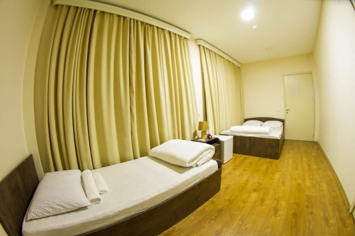 Quarto de hotel com uma cama de solteiro, mesa de cabeceira, abajur e piso de madeira.
