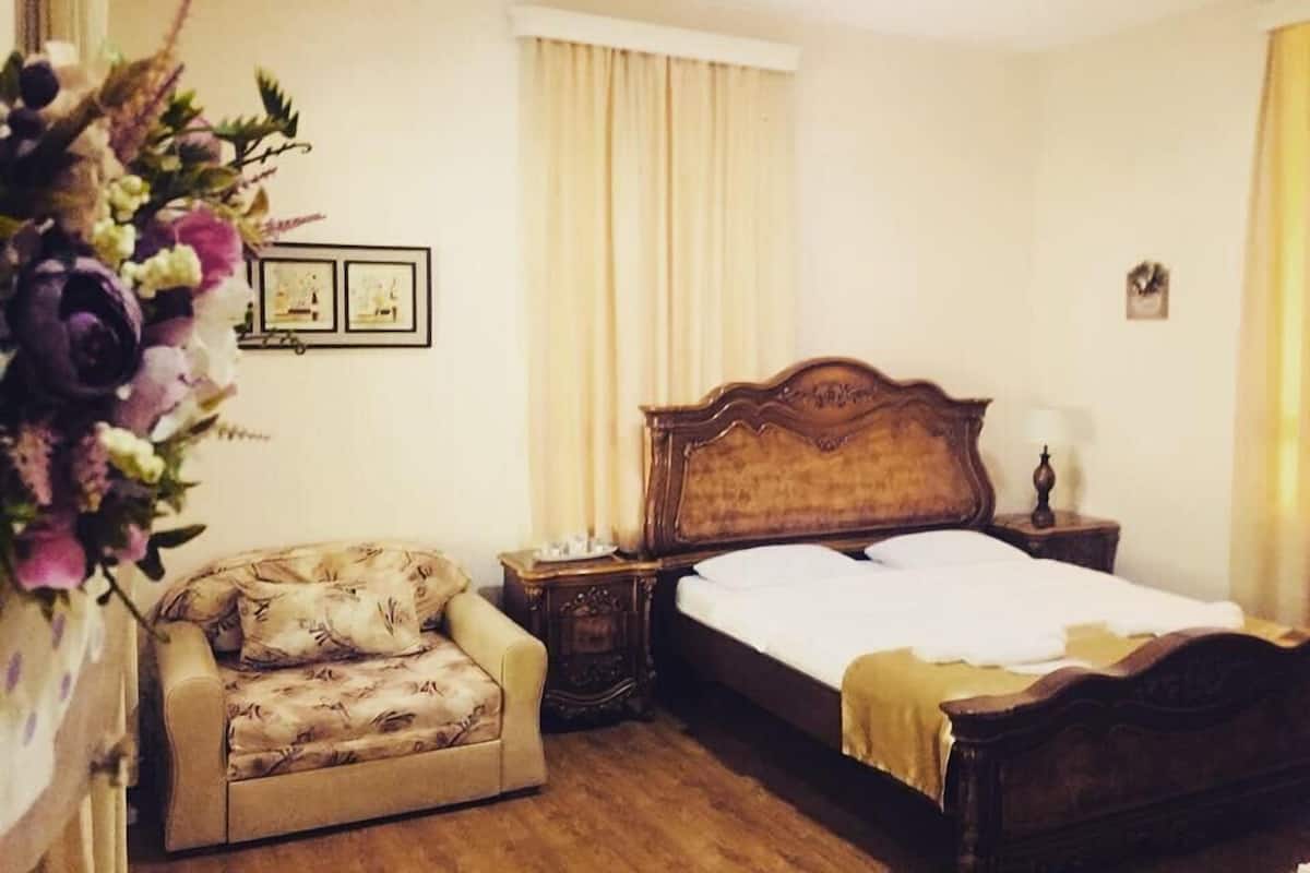 Quarto de hotel com cama de madeira, poltrona estampada, mesa de cabeceira com abajur e janela com cortinas.