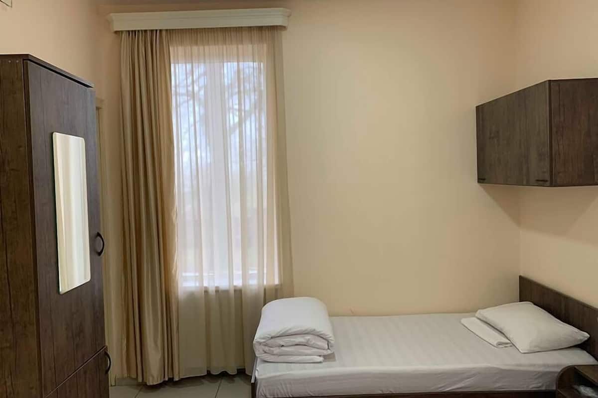 Um quarto pequeno e bem arrumado com uma cama de solteiro, um guarda-roupa de madeira e uma janela com cortinas leves.