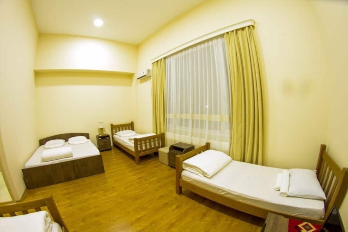 Quarto de hotel com duas camas de solteiro, piso de madeira, janela com cortinas e uma pequena mesa de cabeceira com abajur.