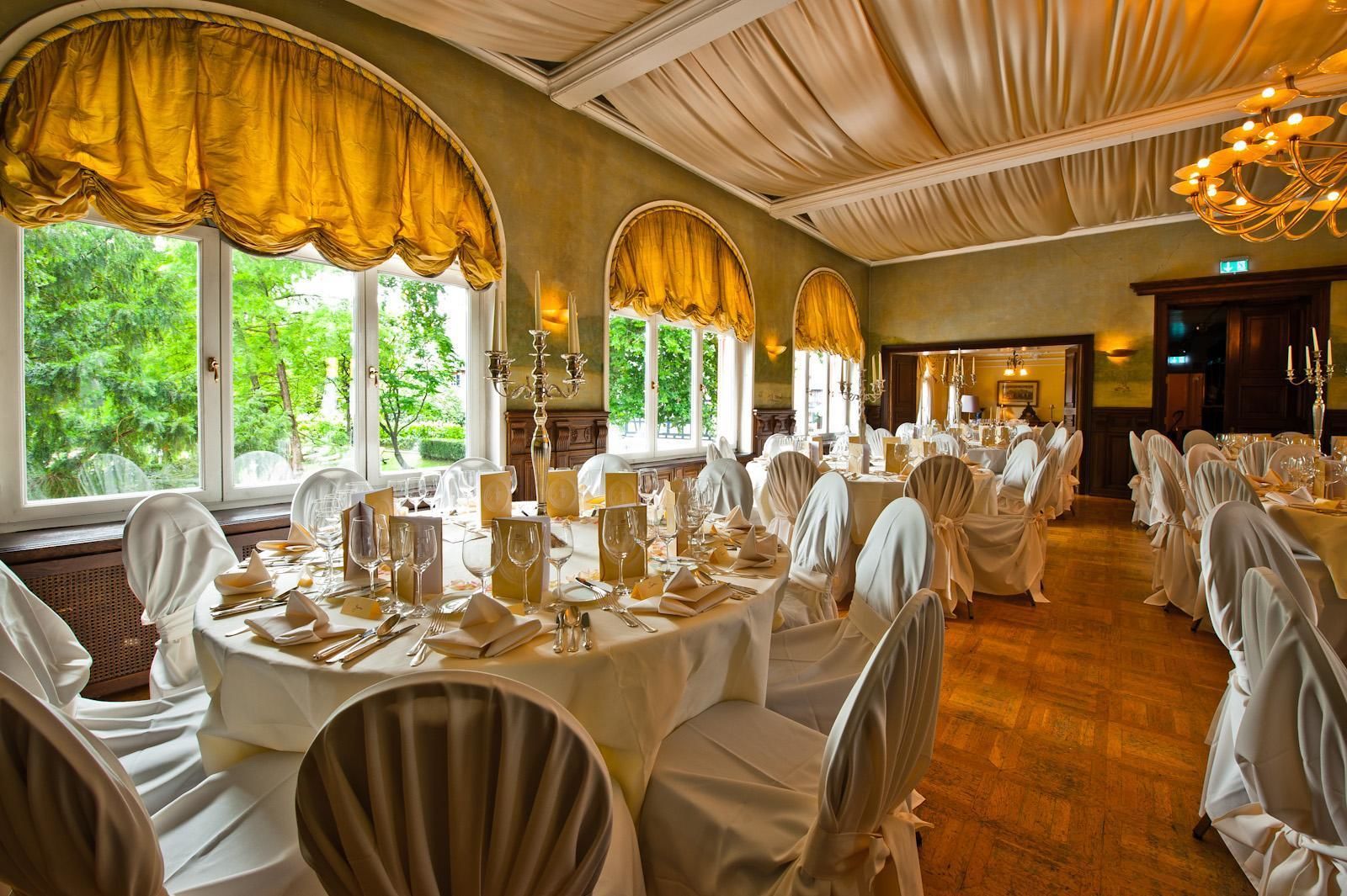 Uma sala de banquetes com mesas redondas preparadas para um evento formal, janelas amplas com cortinas e um candelabro.