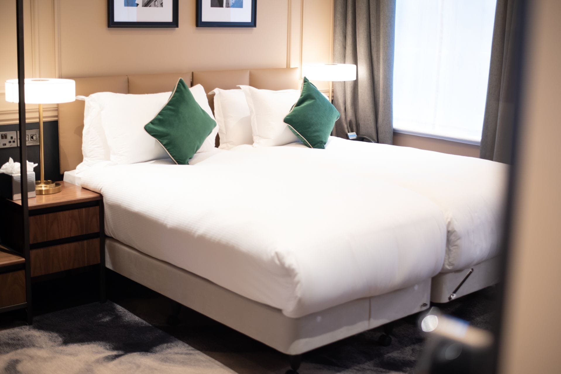 Quarto de hotel com uma cama grande, duas almofadas verdes, uma mesinha de cabeceira com abajur e uma janela com cortinas.