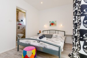 Quarto com cama, um banquinho colorido, banheiro com vaso sanitário e chuveiro.
