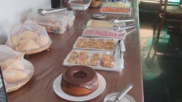 Uma mesa de buffet com diversos alimentos, incluindo bolos, doces e um bolo de chocolate numa placa branca.