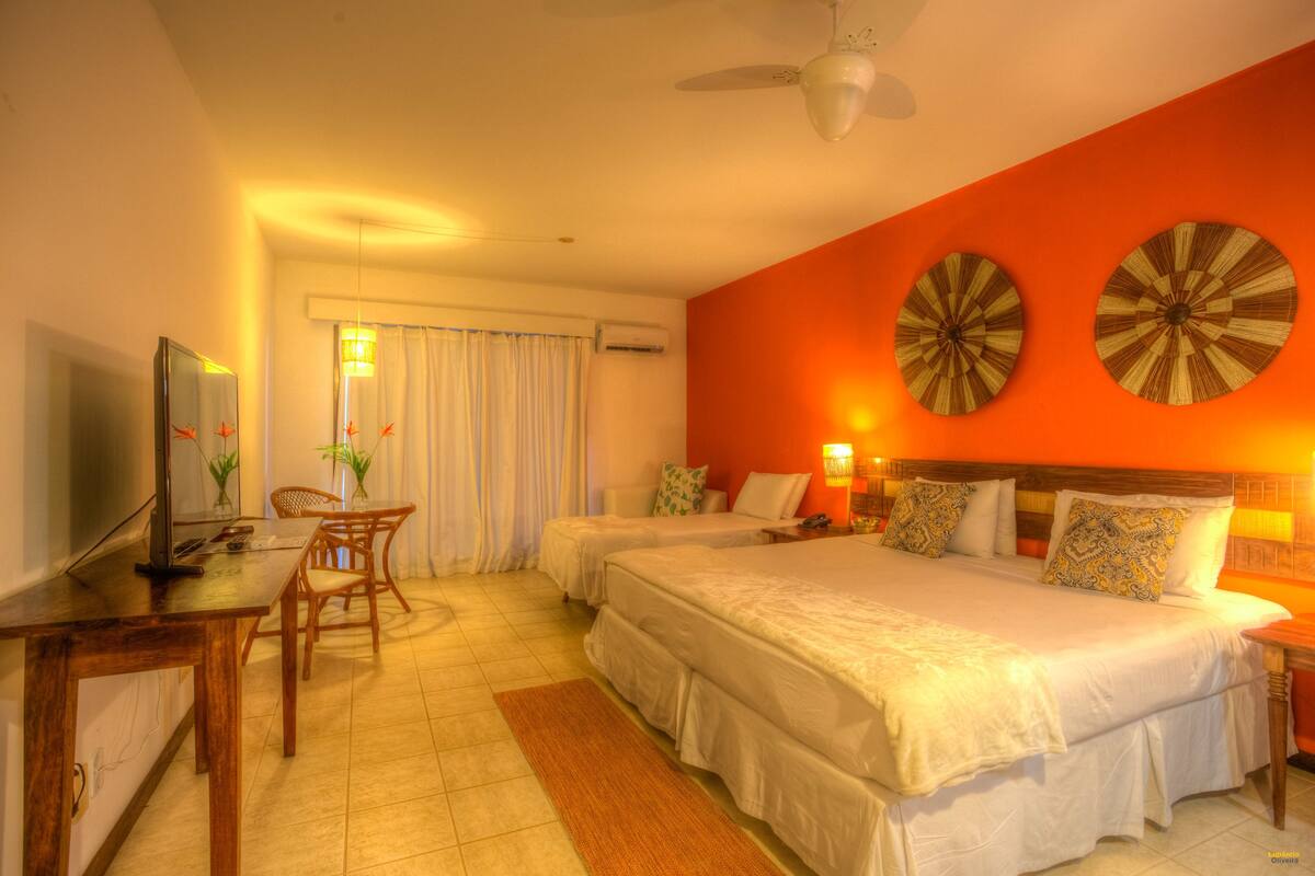 Quarto de hotel com duas camas, uma escrivaninha com televisão, área de refeições com cadeiras e arte decorativa nas paredes.