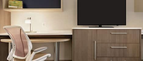 Quarto de hotel com escrivaninha, uma cadeira moderna, televisão e um armário.