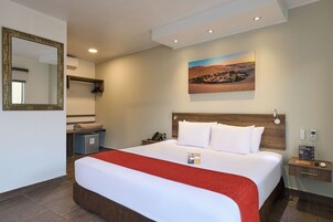 Quarto de hotel com uma cama grande, mesa de cabeceira, espelho, pintura de paisagem desértica e manta vermelha.