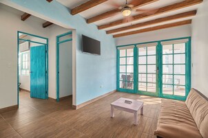 Ampla sala de estar com piso de madeira, um sofá bege, uma mesinha e um ventilador de teto. Há duas portas com molduras azul-turquesa que dão para outros cômodos, e uma grande janela com vista.