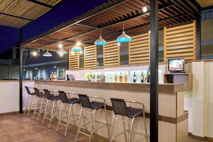 Uma área de bar ao ar livre com cadeiras de metal, balcão com garrafas e luminárias pendentes.