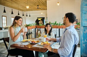 Um grupo de pessoas jantando em uma mesa de madeira em um café com luminárias pendentes e plantas em vasos.