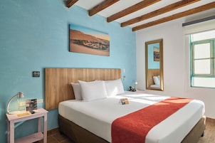 Quarto de hotel com cabeceira de madeira, cama com roupa de cama branca e manta vermelha, mesa de cabeceira com abajur, quadro na parede e janela com vista.