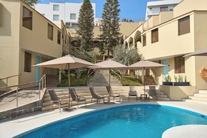 Piscina com espreguiçadeiras e guarda-sóis, cercada por edifícios com escadas que levam a um terraço.