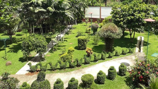 Um jardim bem cuidado com diversos tipos de árvores e arbustos, um caminho pavimentado e uma pequena construção ao fundo.