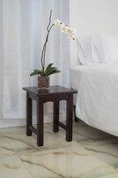 Uma pequena mesa de madeira com uma orquídea em vaso e uma cama branca com cobertura branca ao fundo.