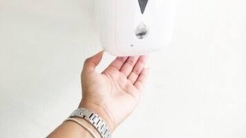 Uma mão segura um dispensador de álcool em gel branco contra uma parede lisa.