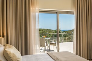 Quarto de hotel com cama, varanda com mesa e cadeiras, e vista para o mar e vegetação.