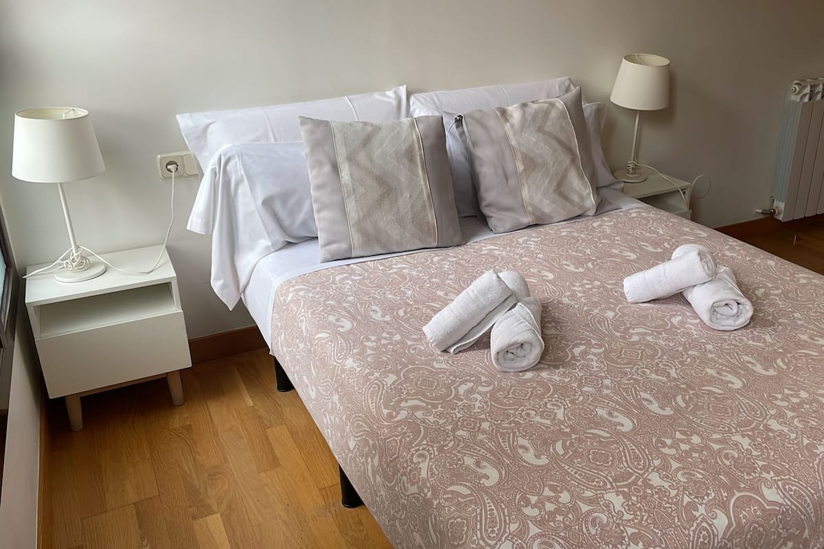 Cama bem arrumada com colcha estampada, dois toalhas enroladas e duas luminárias de cabeceira em mesas de cabeceira brancas.