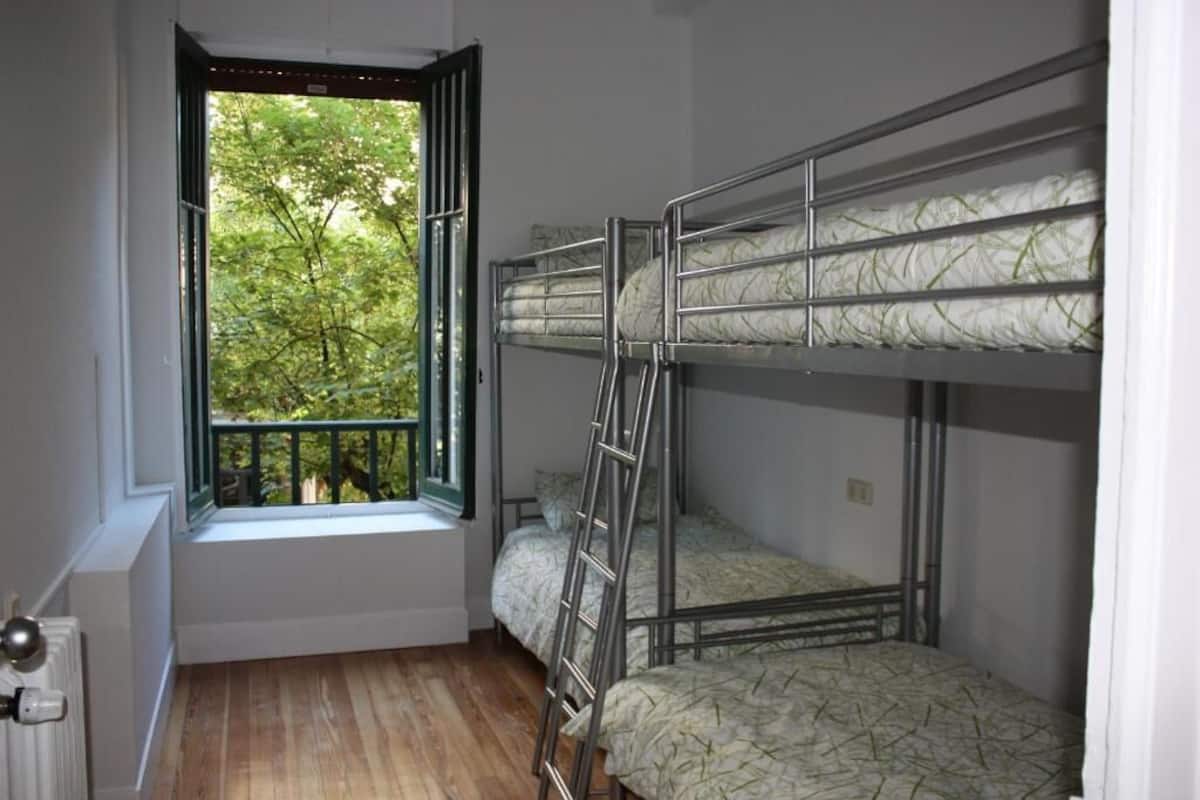 Quarto pequeno com beliches, janela com vista para árvores e piso de madeira.