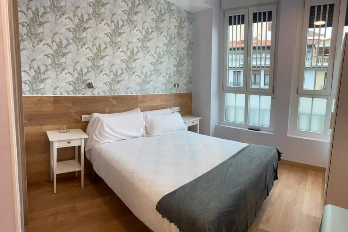 Quarto com cabeceira de madeira, cama branca, manta cinza, mesa de cabeceira com um copo e janela com vista para edifícios.