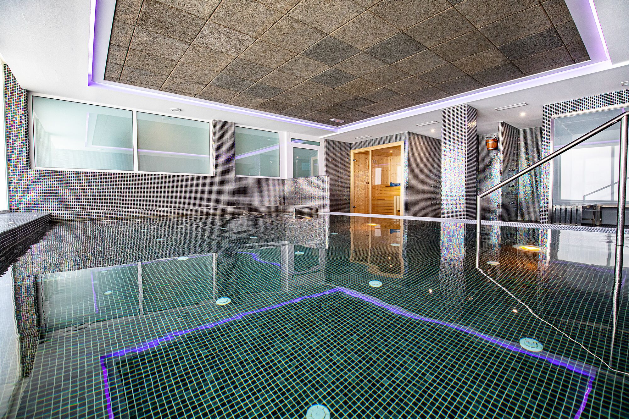 Piscina interior moderna com piso de vidro, rodeada por paredes com azulejos e uma porta de madeira para um vestiário.
