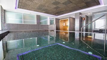 Piscina interior moderna com piso de vidro, rodeada por paredes com azulejos e uma porta de madeira para um vestiário.