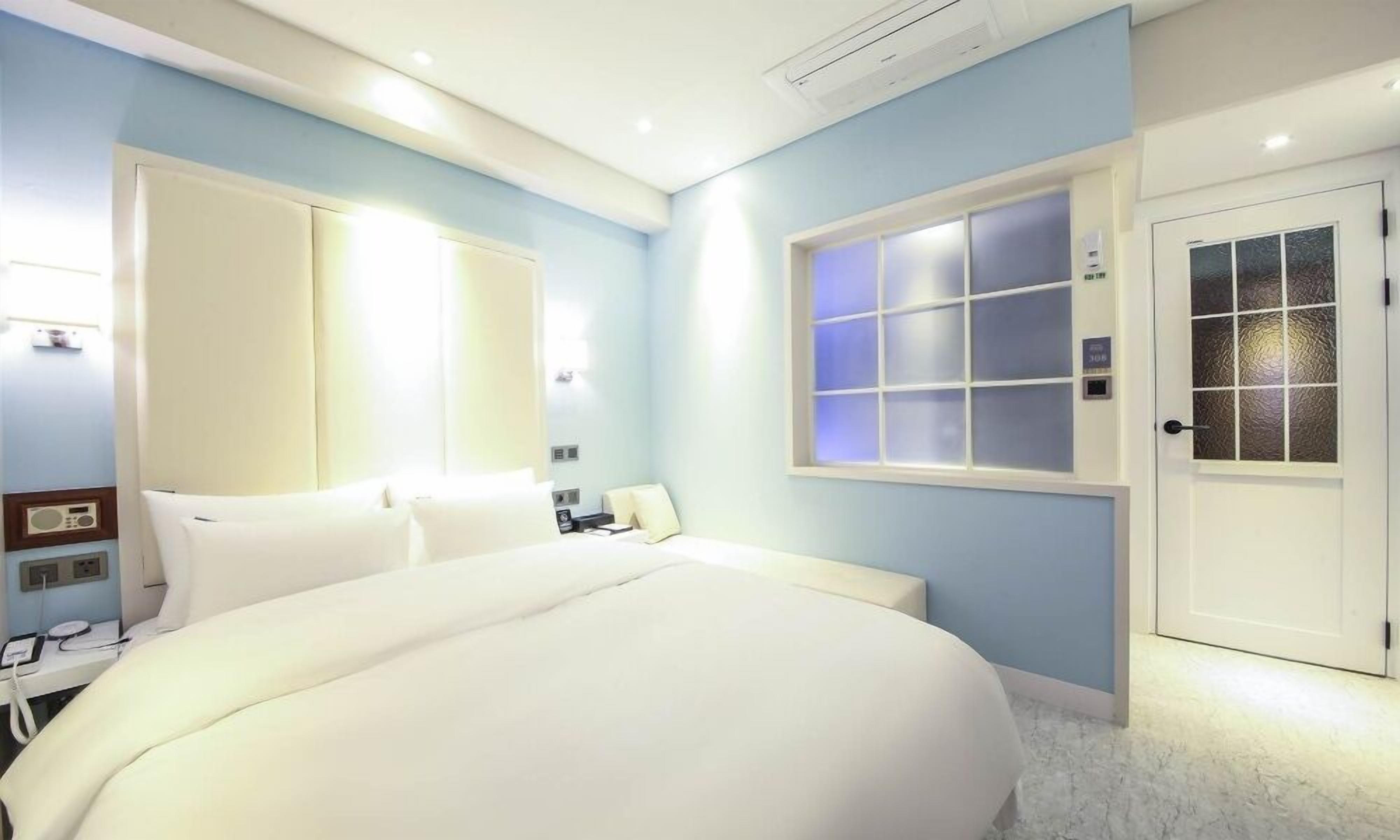 Quarto de hotel com uma cama grande, mesas de cabeceira, um rádio, um telefone, uma janela com vidro jateado e uma porta com painel de vidro jateado.