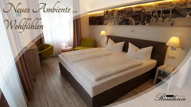 Quarto de hotel com uma cama grande, duas mesas de cabeceira com abajures, uma cadeira verde e um quadro pendurado na parede.
