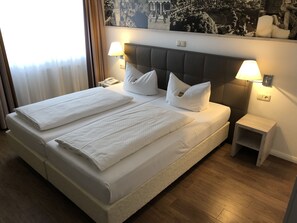 Uma cama de casal com roupa de cama branca, duas mesas de cabeceira com abajures, um telefone e um interruptor de luz na parede.
