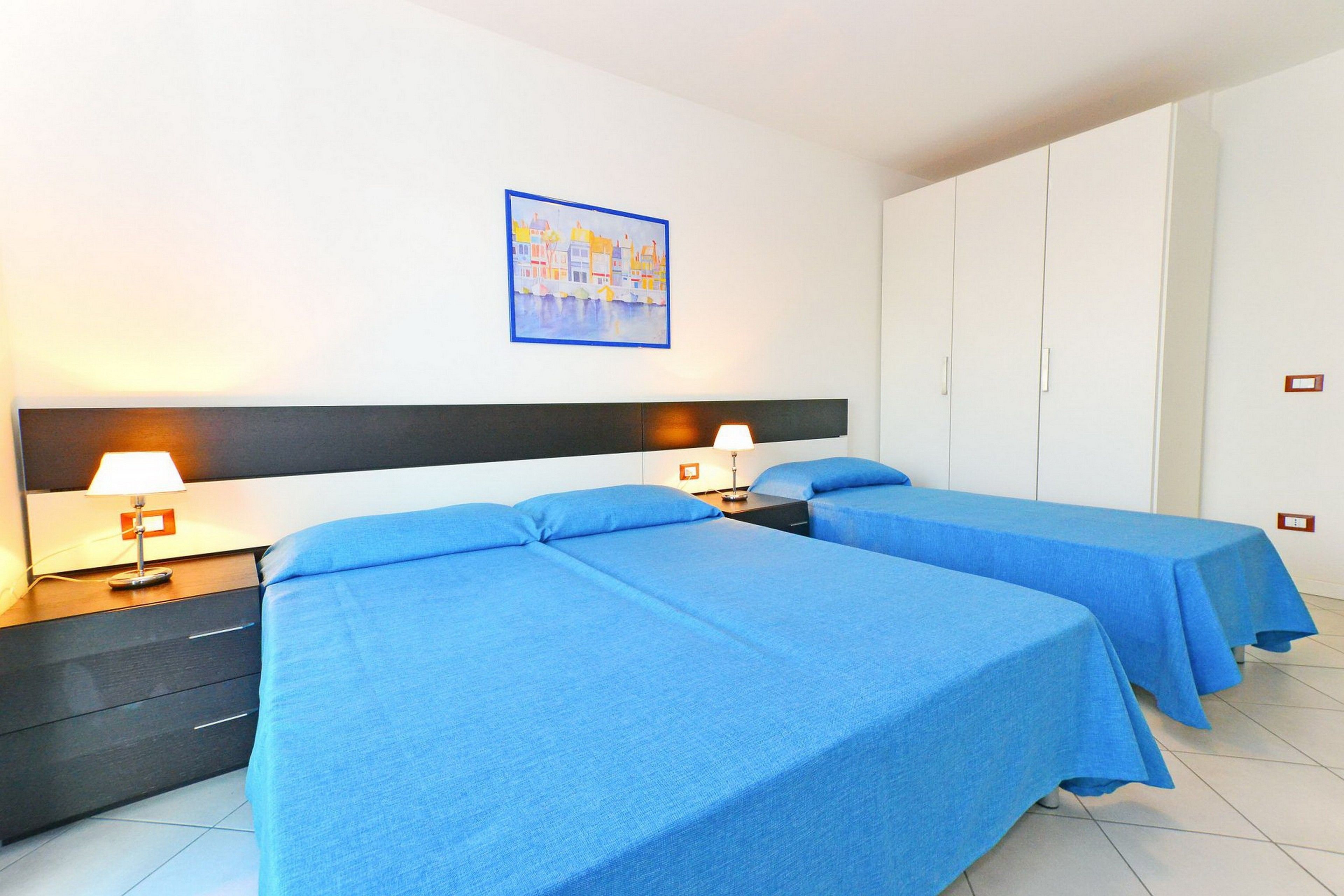 Quarto de hotel com duas camas de solteiro, mesa de cabeceira com abajur, cômoda e guarda-roupa.
