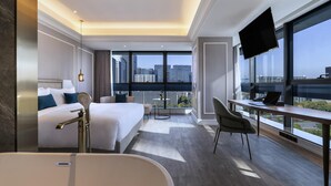 Quarto de hotel moderno com uma cama grande, uma mesa com um laptop, uma TV e uma varanda com vista para a cidade.