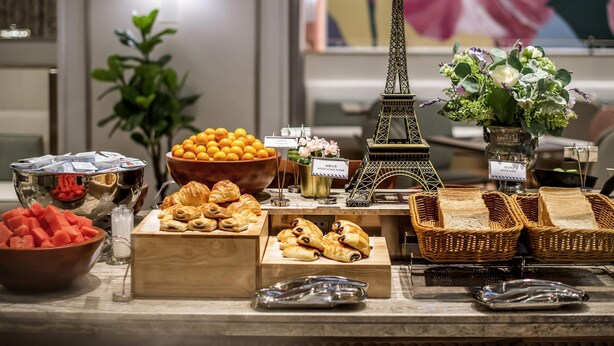 Uma mesa de buffet com diversos alimentos, incluindo croissants, doces e uma pequena réplica da Torre Eiffel.