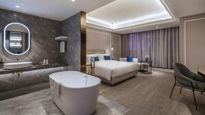 Quarto de hotel moderno com uma banheira grande, cama de casal, área de estar e uma janela ampla com cortinas.