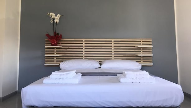 Quarto de hotel com cama, roupa de cama branca, cabeceira de madeira e um vaso com flores brancas.