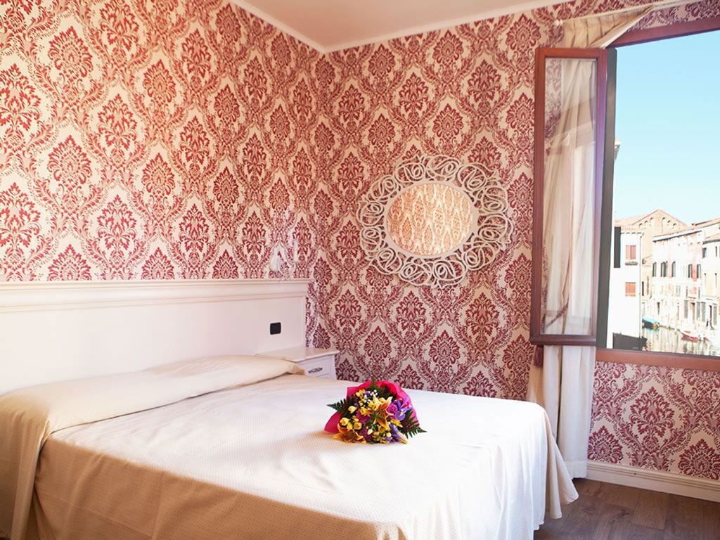 Quarto de hotel com papel de parede estampado, uma cama com um arranjo de flores e vista para edifícios pela janela.