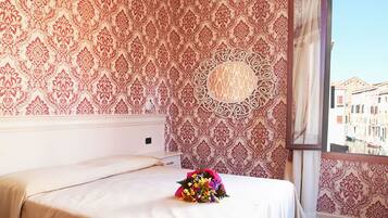Quarto de hotel com papel de parede estampado, uma cama com um arranjo de flores e vista para edifícios pela janela.
