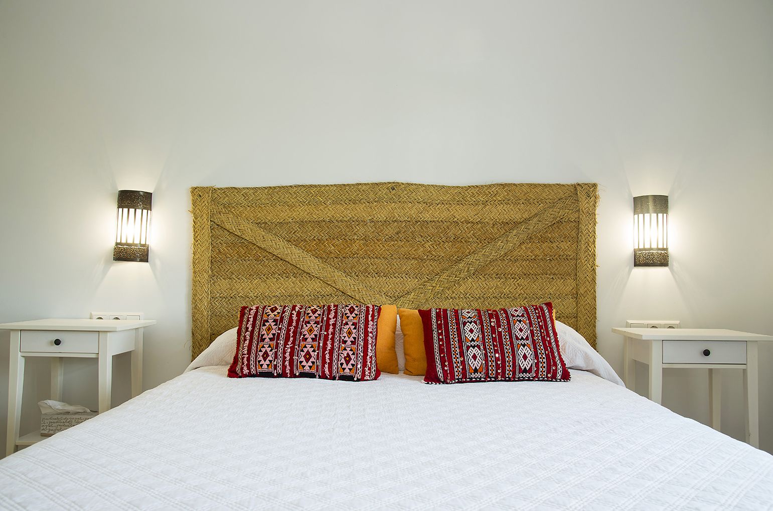 Uma cama com colcha branca, duas almofadas estampadas e uma cabeceira trançada. Há duas mesas de cabeceira com luminárias de cada lado da cama.