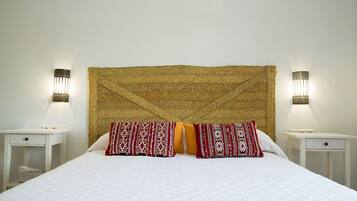 Uma cama com colcha branca, duas almofadas estampadas e uma cabeceira trançada. Há duas mesas de cabeceira com luminárias de cada lado da cama.