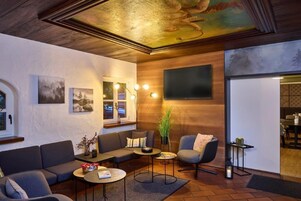 Uma sala de estar moderna com teto de madeira, uma grande TV de tela plana e uma área de estar aconchegante com sofá e poltronas.