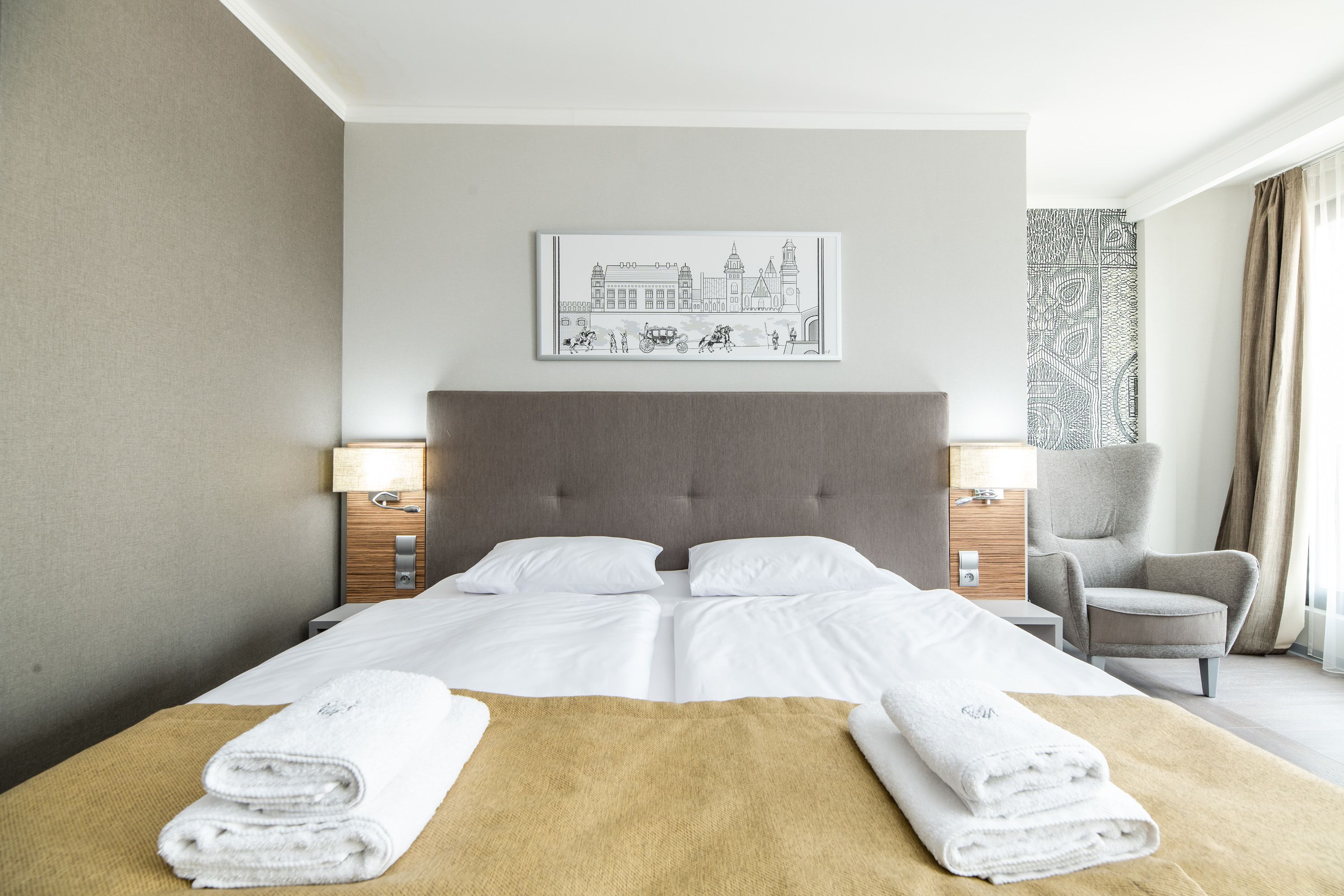Quarto de hotel com uma cama grande, duas mesas de cabeceira, uma cadeira e uma pintura com vista urbana na parede.