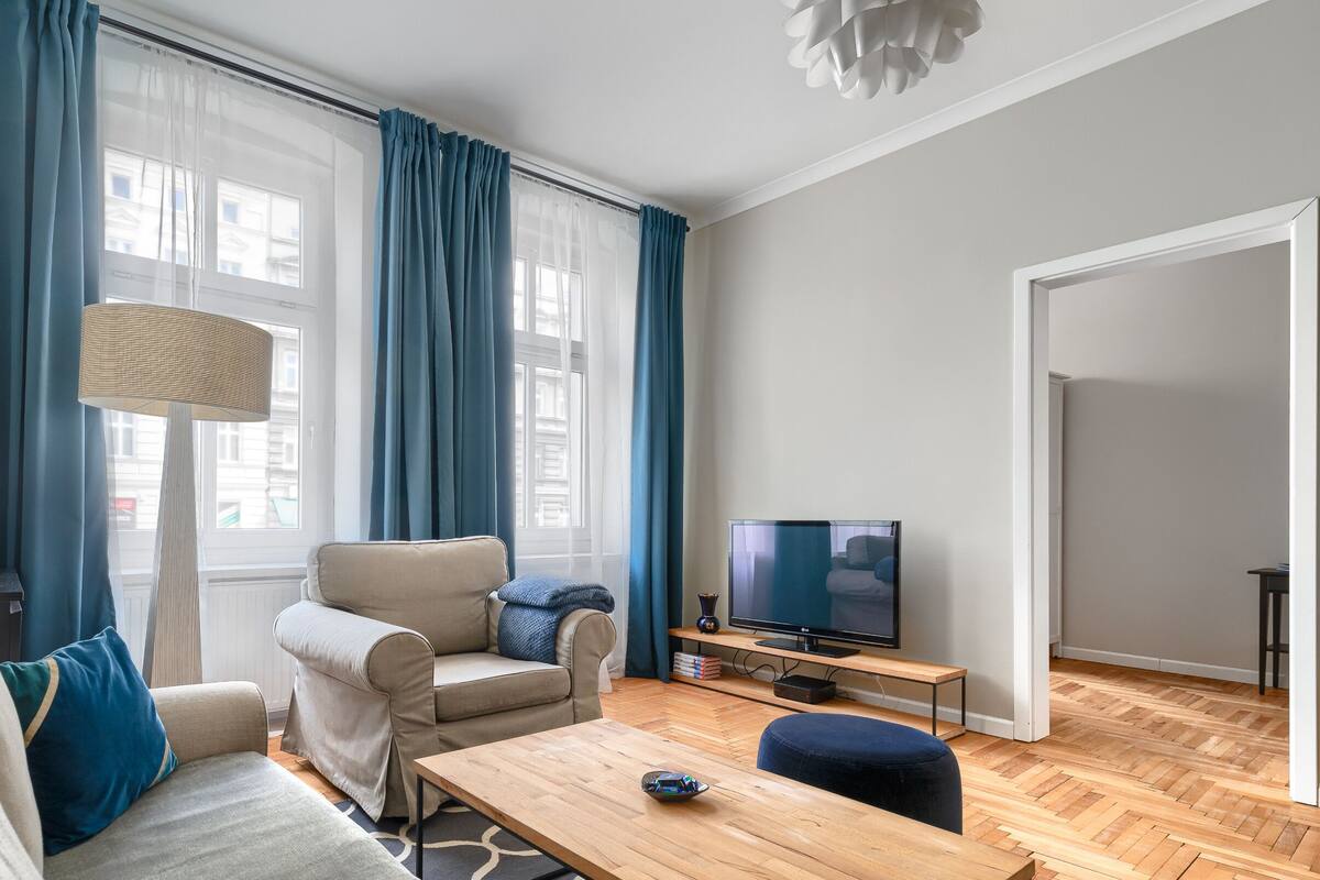 Uma sala de estar moderna com um sofá, poltrona e um banquinho, uma mesa de centro de madeira, uma TV em um móvel, e uma janela grande com cortinas.