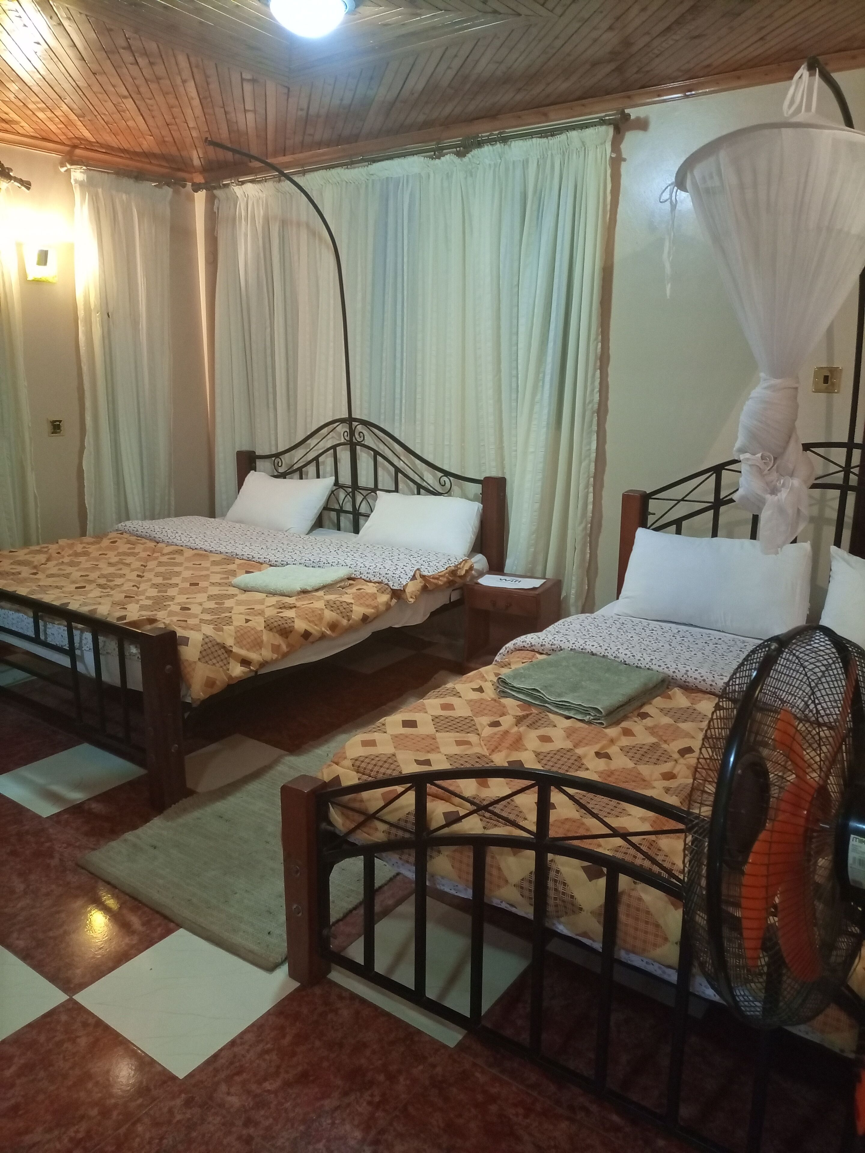 Quarto com duas camas, cada uma com estrutura de madeira e roupa de cama estampada. Há um ventilador e uma mosquiteira pendurada sobre uma das camas.